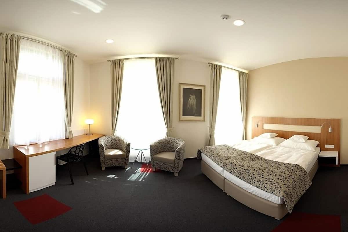 Quarto de hotel com cama, escrivaninha, duas cadeiras, uma mesa pequena e um quadro na parede.
