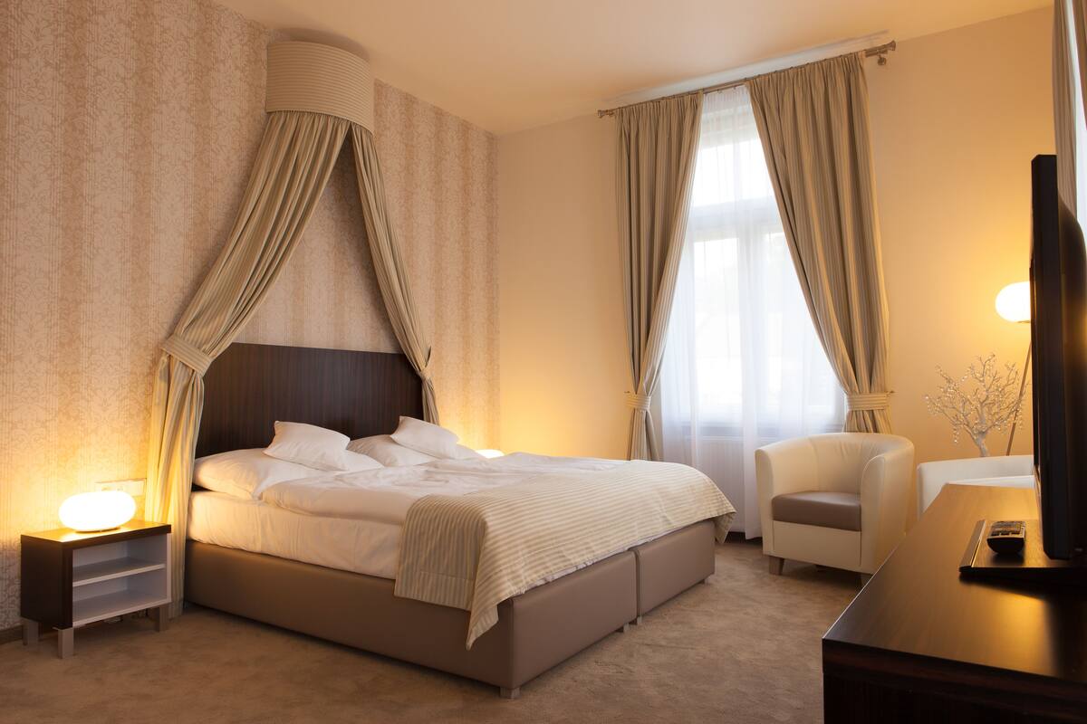 Quarto de hotel com cama grande, mesa de cabeceira com abajur, cadeira e televisão.
