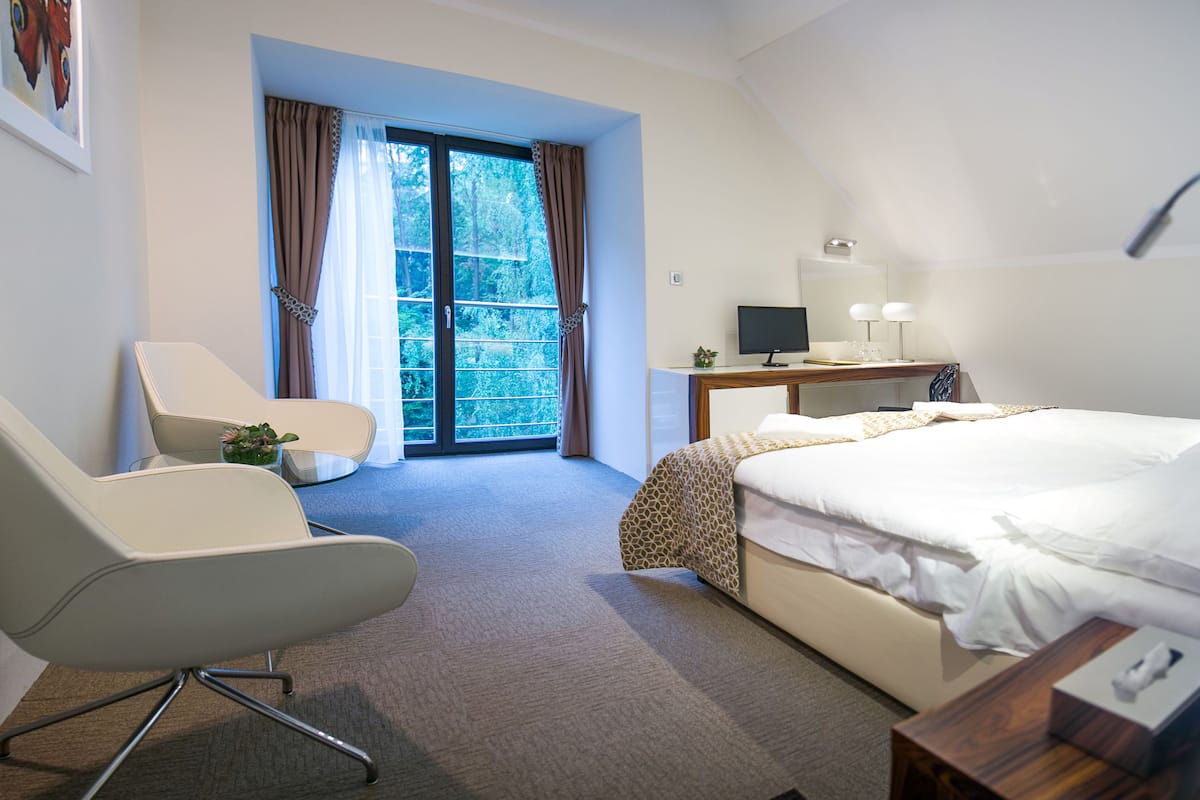 Quarto de hotel com uma cama, uma escrivaninha com computador, uma cadeira e vista para o exterior através de uma porta de vidro.
