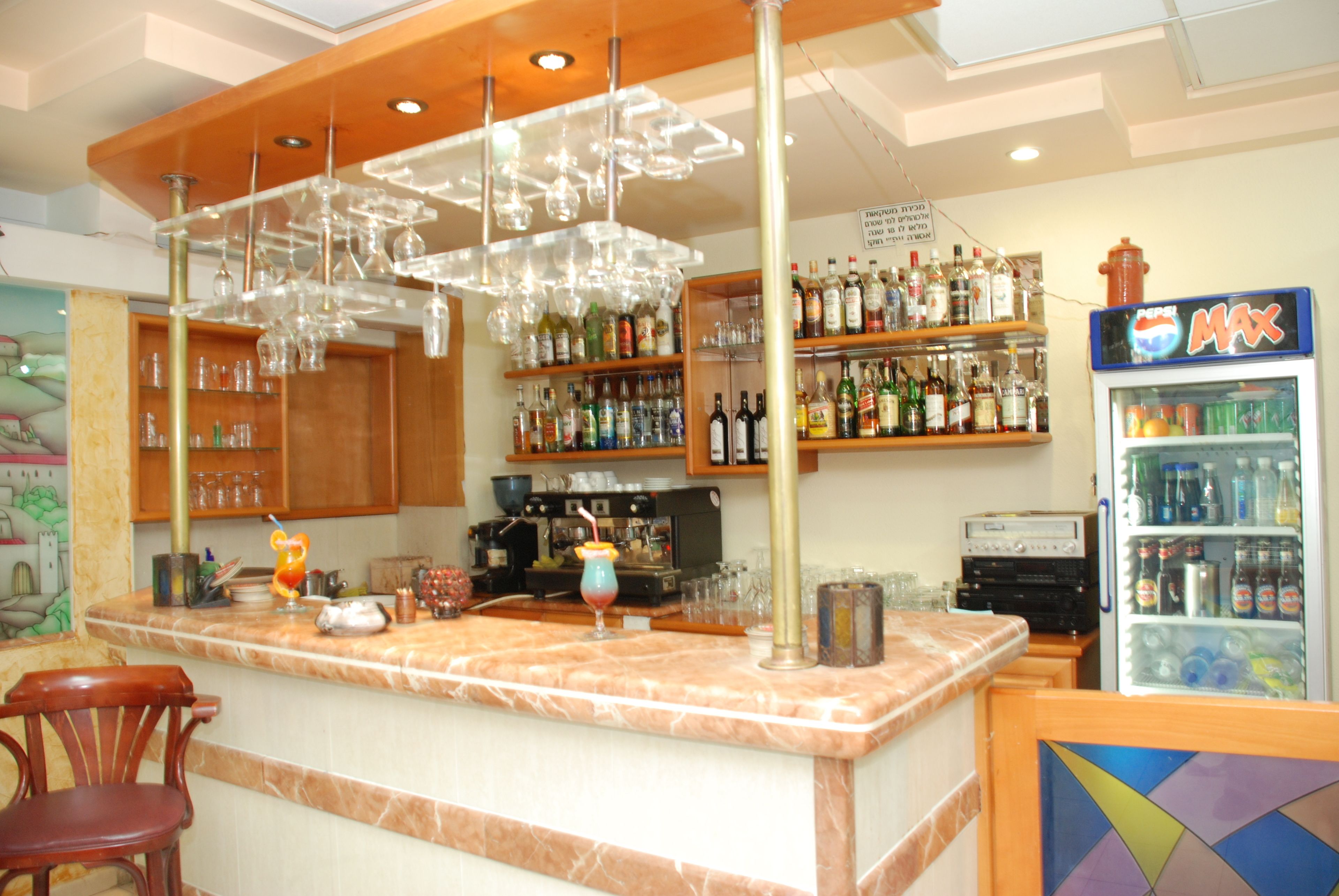 Uma área de bar com bancada de mármore, lustres de vidro pendurados, prateleiras com garrafas de bebidas e um refrigerador de Pepsi Max.