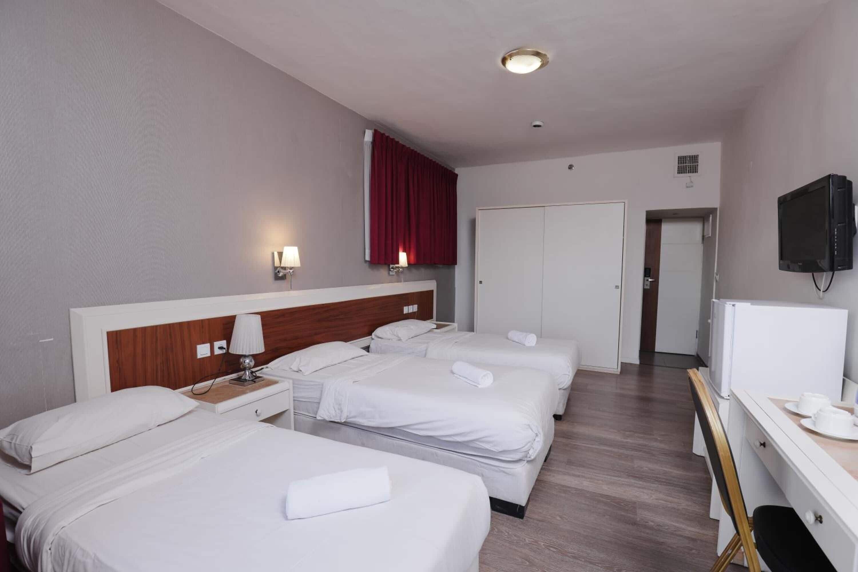 Quarto de hotel com três camas, uma secretária com cadeira, televisão e porta para um banheiro.
