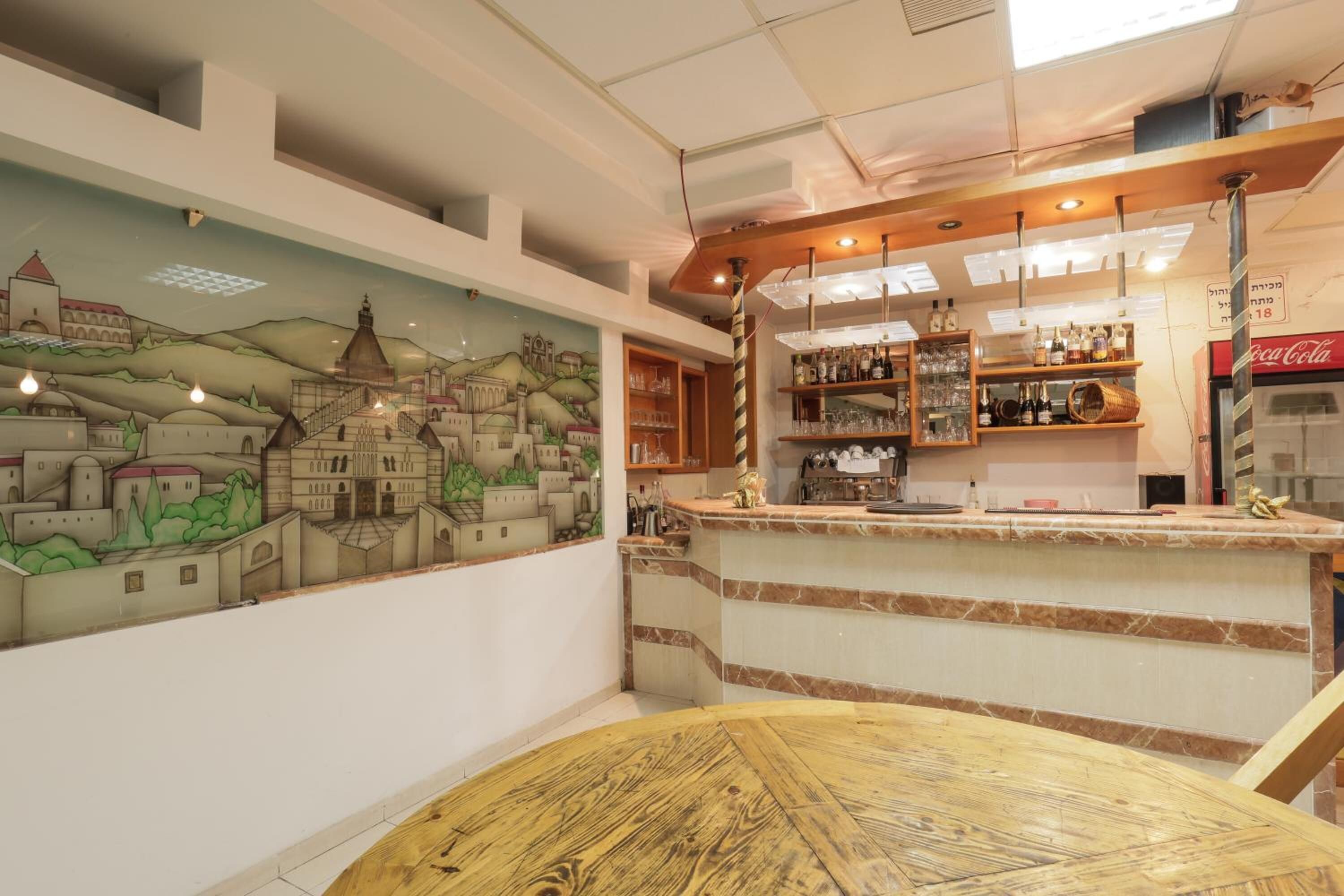 Uma área de bar com um mural de uma cidade, prateleiras com garrafas e uma geladeira Coca-Cola.