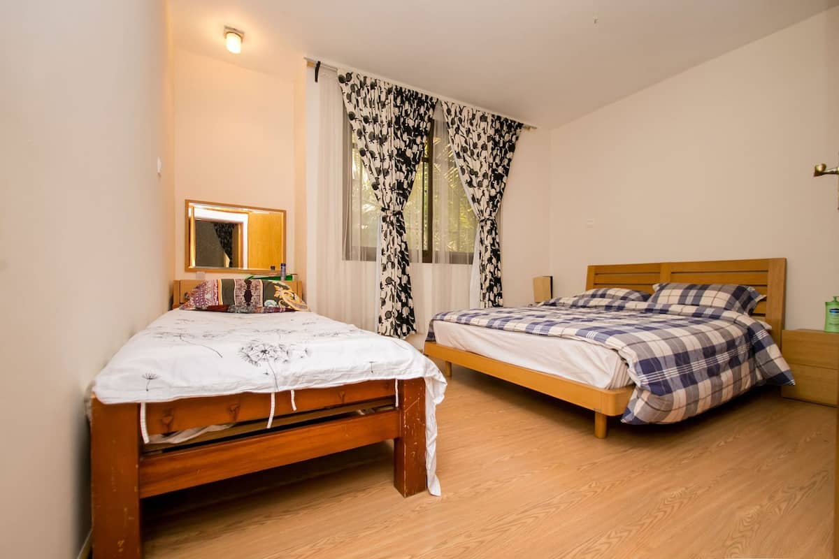 Quarto com duas camas de madeira, uma mesa de cabeceira, um espelho e uma janela com cortinas estampadas.