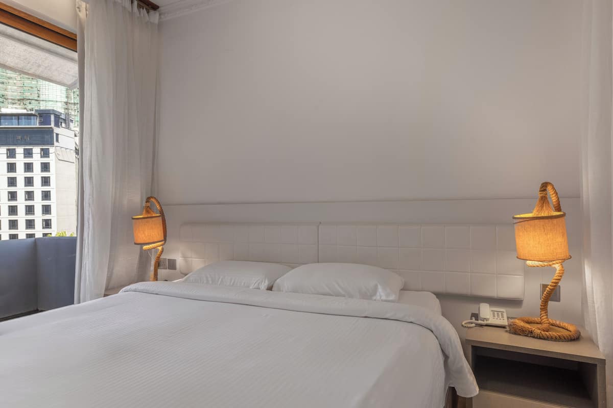 Quarto de hotel com uma cama grande, roupa de cama branca, duas luminárias de cabeceira, um criado-mudo e uma janela com cortinas.