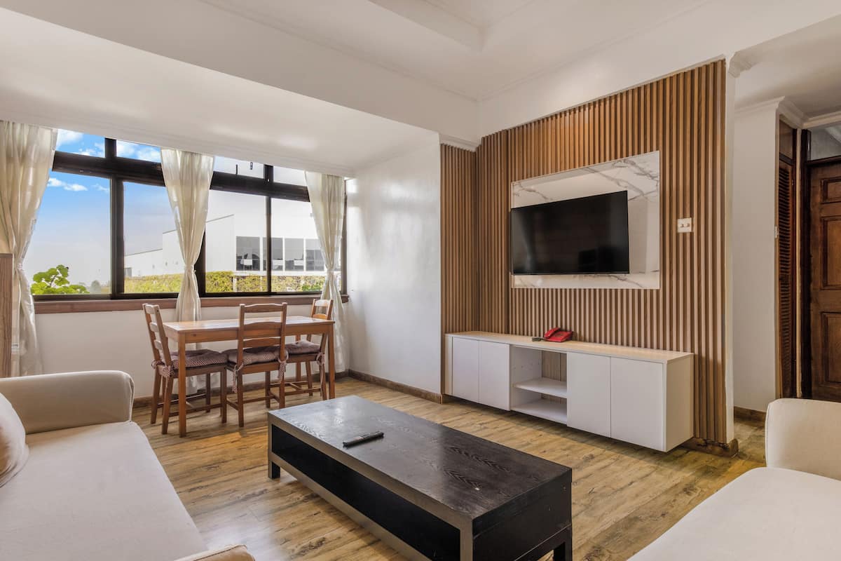 Uma sala de estar moderna com piso de madeira, uma TV fixada na parede, uma área de jantar com mesa e cadeiras, e um sofá.