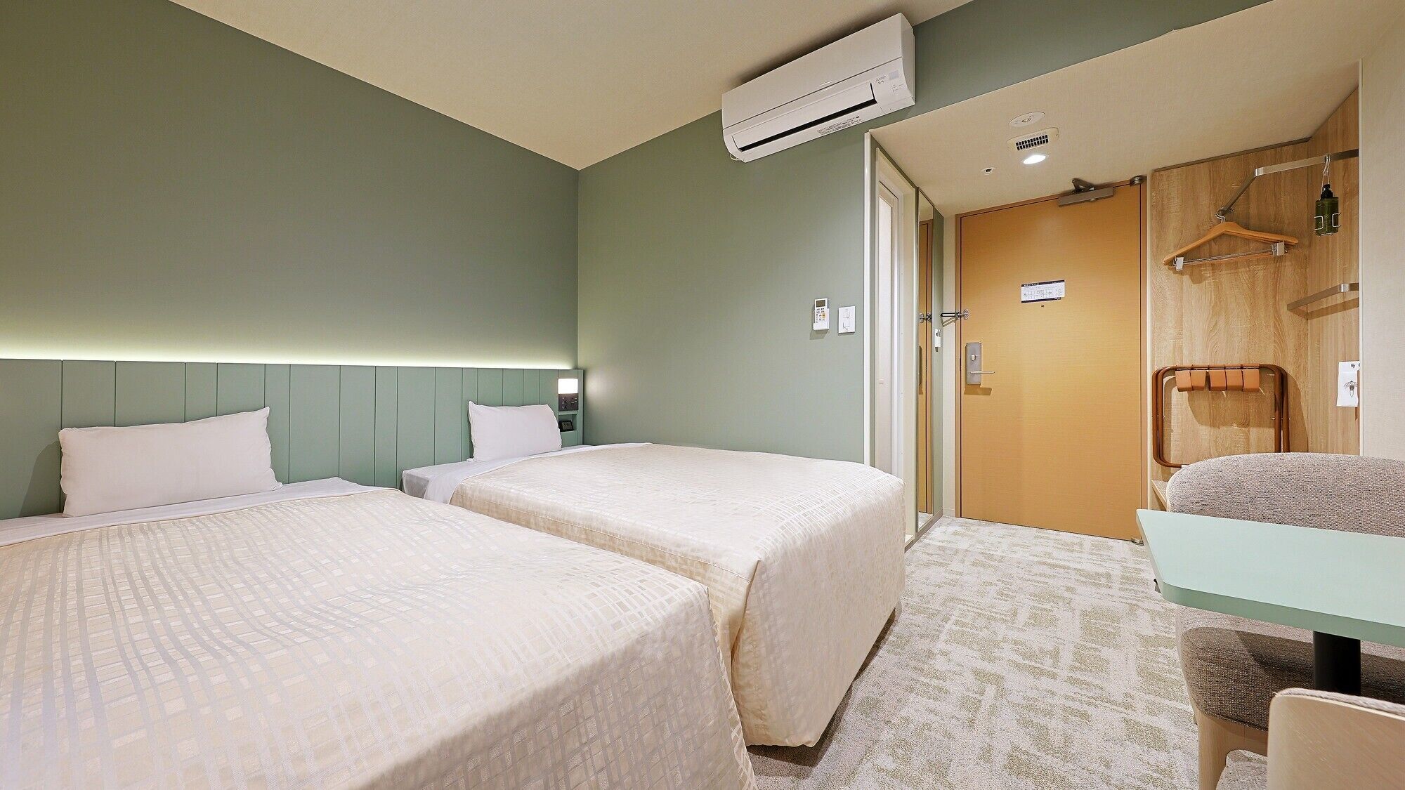 Quarto de hotel com duas camas, uma escrivaninha, uma cadeira, aparelho de ar condicionado e uma porta.