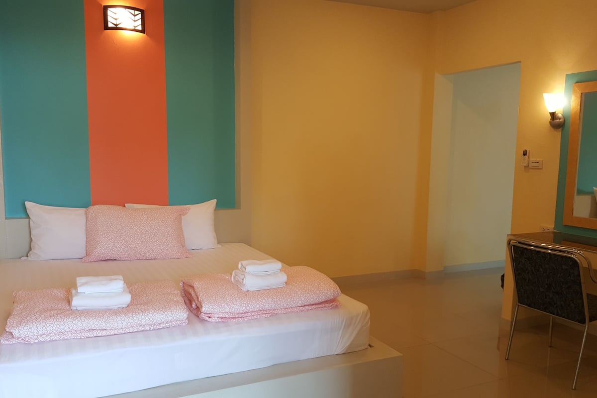 Quarto de hotel com cama, almofadas, toalha dobrada, cadeira, espelho e luminária fixada na parede.