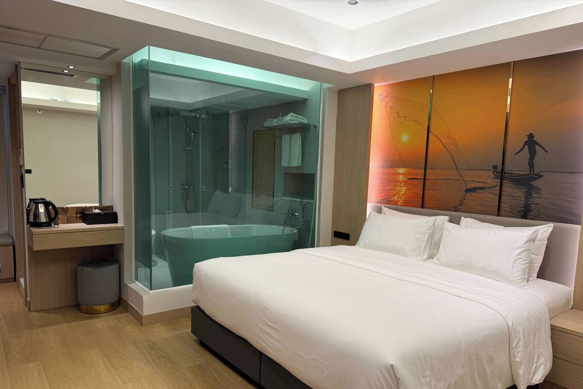 Quarto de hotel moderno com uma cama grande, chuveiro em cabine de vidro, escrivaninha com cafeteira e um mural com o tema do pôr do sol na parede.