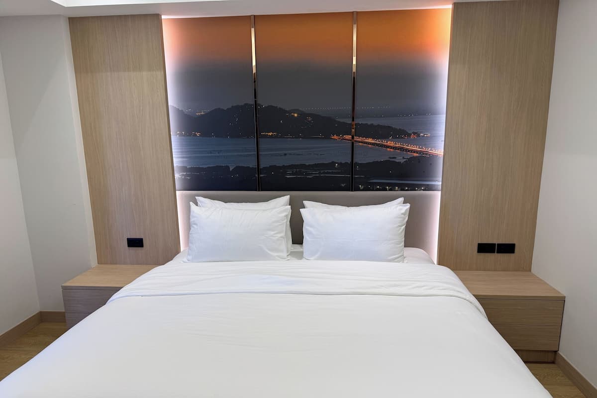 Quarto de hotel com uma cama grande, duas almofadas, cabeceira de madeira e vista noturna de um horizonte urbano com uma ponte.
