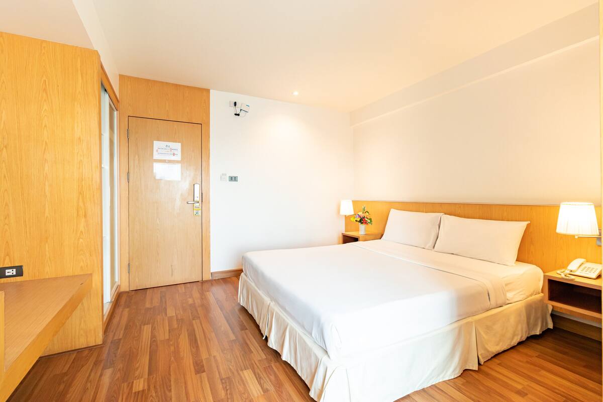 Quarto de hotel com piso de madeira, uma cama grande com roupa de cama branca, uma mesinha de cabeceira com abajur e uma porta com um aviso.