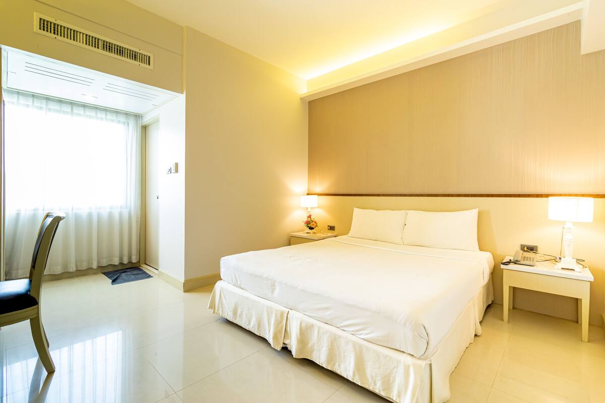 Quarto de hotel com uma cama grande, mesa de cabeceira com abajur, uma cadeira e janela com cortinas.