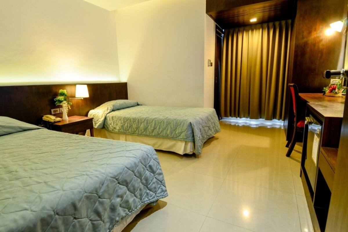 Quarto de hotel com duas camas, uma escrivaninha, uma cadeira, um abajur e uma cortina.