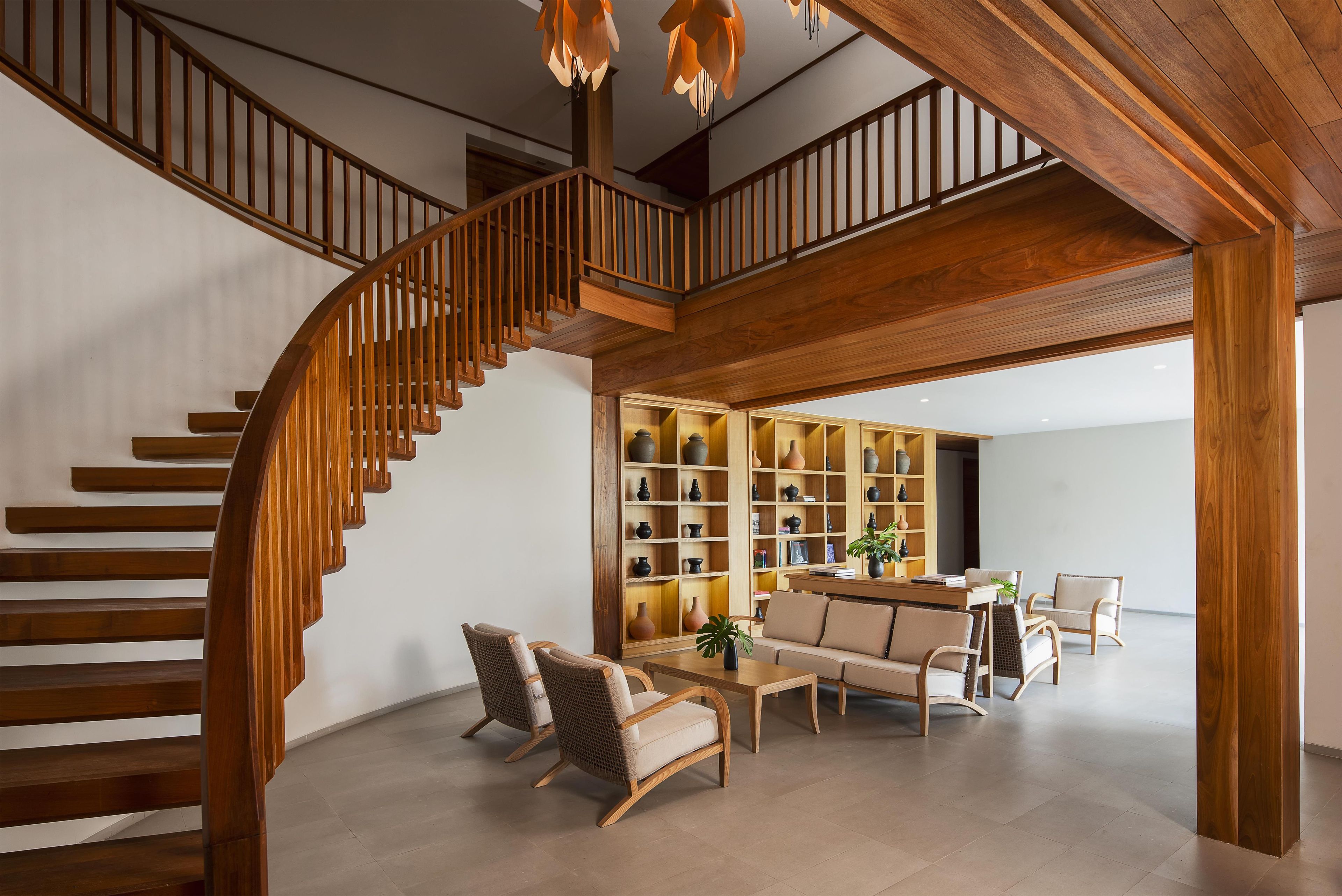 Amplo interior com escada de madeira, área de estar com sofás e poltronas, e estante repleta de objetos decorativos.
