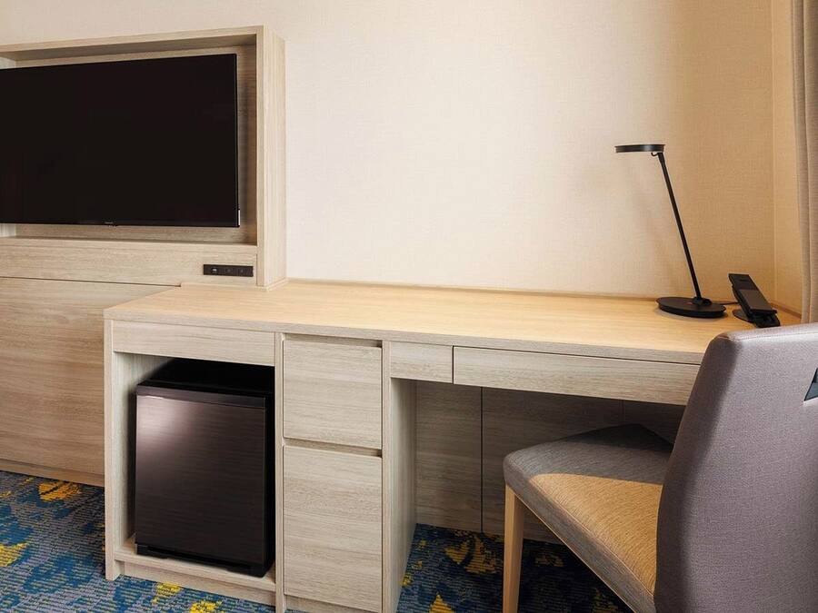 Quarto de hotel com escrivaninha de madeira, uma cadeira, televisão e abajur.
