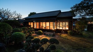 Uma casa japonesa tradicional com telhado de telhas, janelas amplas e um jardim bem cuidado com diversos arbustos e um caminho de pedra.