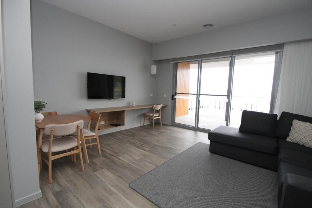 Uma sala de estar moderna com televisão de tela plana, um sofá escuro, uma mesa de jantar com cadeiras e uma grande porta de vidro deslizante que dá para uma varanda.