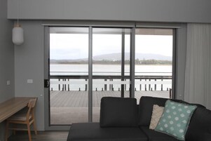 Sala de estar moderna com sofá preto, mesa de madeira e vista para um corpo de água através de grandes portas de correr de vidro.