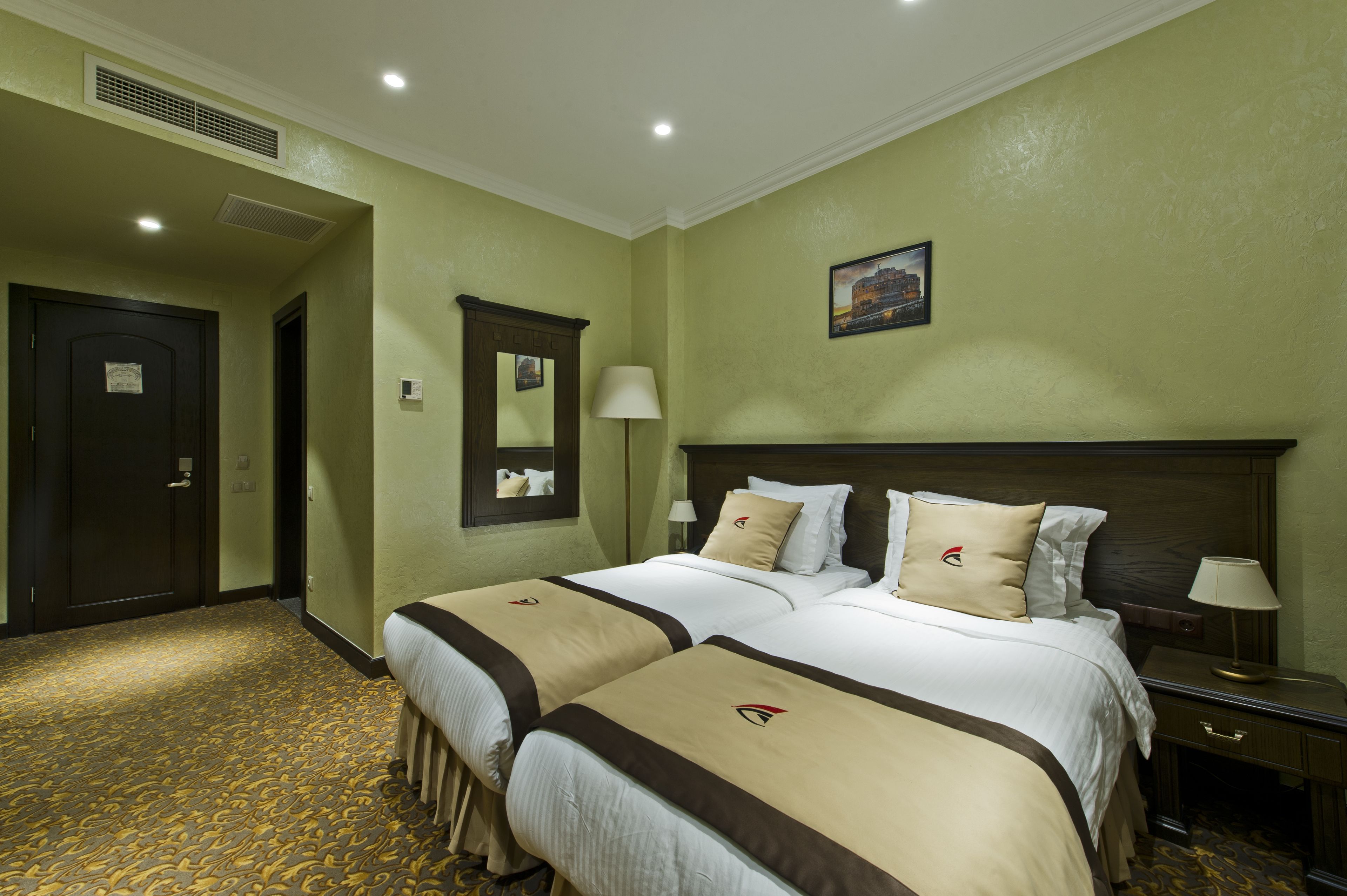 Quarto de hotel com duas camas, uma mesa de cabeceira, uma luminária, um espelho e uma porta.