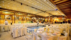 Uma sala de banquetes com mesas redondas preparadas para um evento formal, área de piscina e teto alto com vigas aparentes.