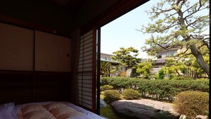 Quarto japonês tradicional com piso de tatame, cama com roupa de cama branca e vista para um jardim com pedras e árvores.