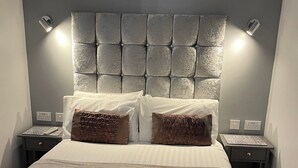 Quarto de hotel com cabeceira acolchoada, duas mesas de cabeceira com luminárias, cama com manta texturizada e dois almofados estampados.