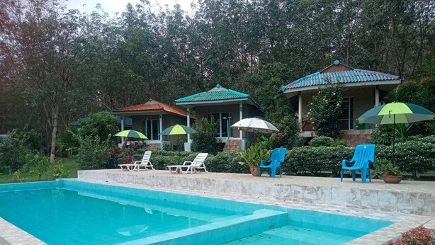 Piscina com espreguiçadeiras e guarda-sóis, cercada por vegetação exuberante e edifícios com telhados em cerâmica.