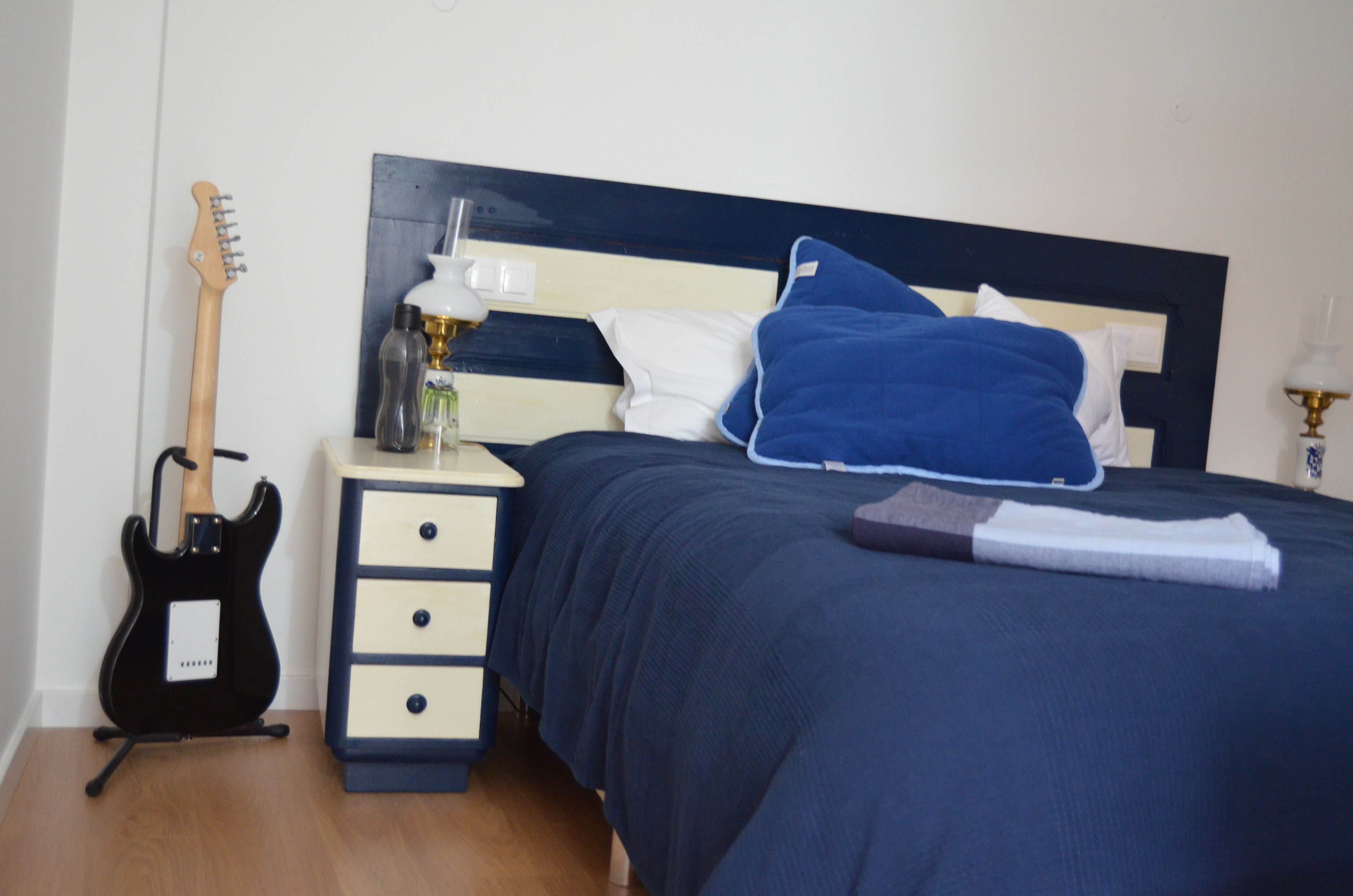Quarto com cama, mesa de cabeceira, abajur e um violão.