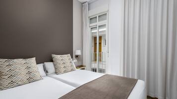 Quarto de hotel com duas camas, mesa de cabeceira, luminária e vista para o exterior através de uma porta.