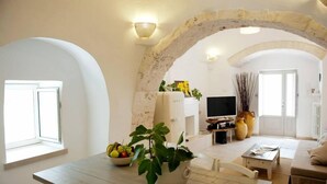 Uma sala de estar ampla com um arco de pedra, um sofá branco, uma mesa de madeira com uma tigela de frutas e uma televisão.