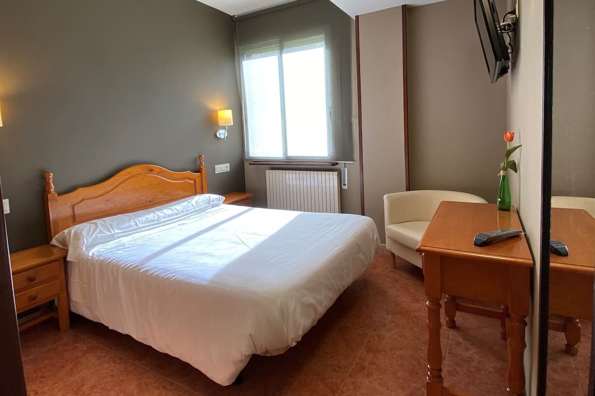 Quarto de hotel com uma cama grande, mesa de cabeceira, cadeira, escrivaninha, radiador e janela com persianas.