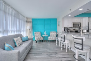 Uma sala de estar moderna com um sofá cinzento, duas poltronas estampadas, uma mesa redonda pequena e uma área de cozinha com armários brancos e eletrodomésticos de aço inoxidável.
