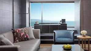 Uma sala de estar moderna com um sofá, uma poltrona azul e uma mesa de centro com maçãs. Uma janela grande oferece vista para o mar e colinas distantes.