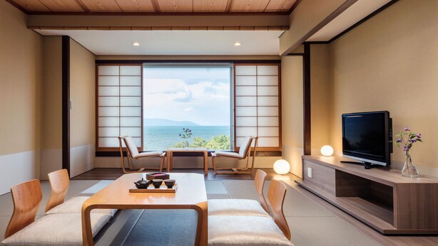 Quarto moderno com tatames, uma mesa de madeira, cadeiras, televisão e vista para o mar.