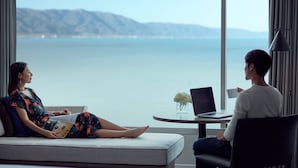 Quarto com uma grande janela que oferece vista para um corpo de água e montanhas. Há um sofá e uma poltrona com um laptop sobre uma mesinha.