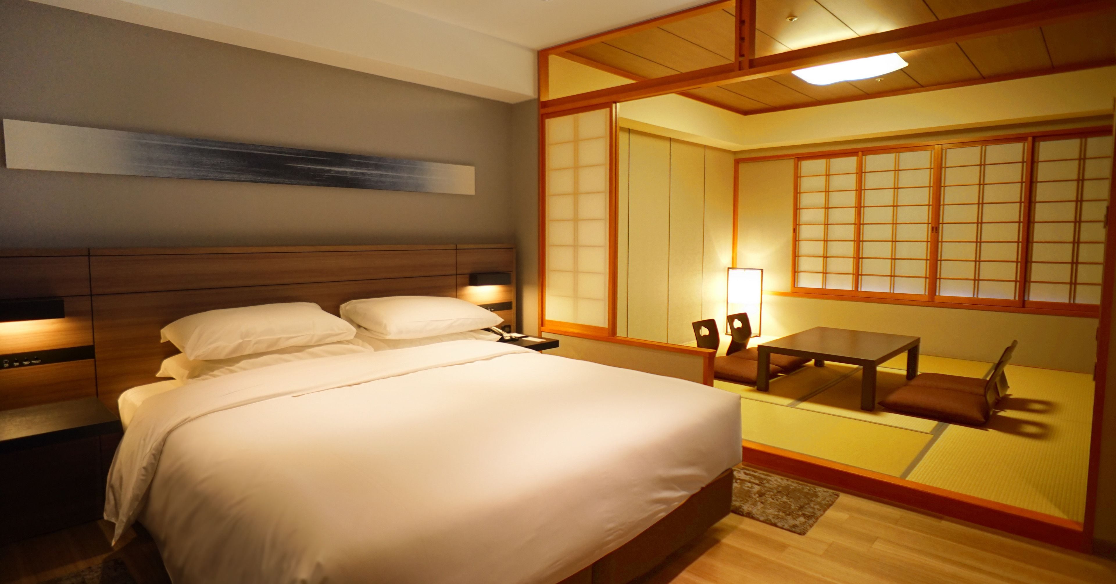 Quarto de hotel com uma cama grande, mesa de cabeceira, abajur e portas japonesas tradicionais.