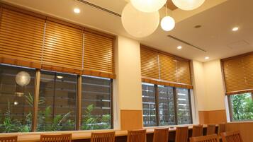 Sala de conferências com mesas e cadeiras de madeira, janelas amplas com persianas e luminárias pendentes.