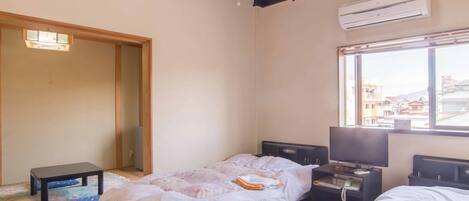 Quarto com cama, escrivaninha, cadeira, televisão e janela com vista para prédios.