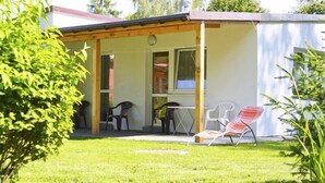Um alpendre coberto com mesa e cadeiras, uma pequena área de jardim e um edifício com fachada branca e telhado vermelho.