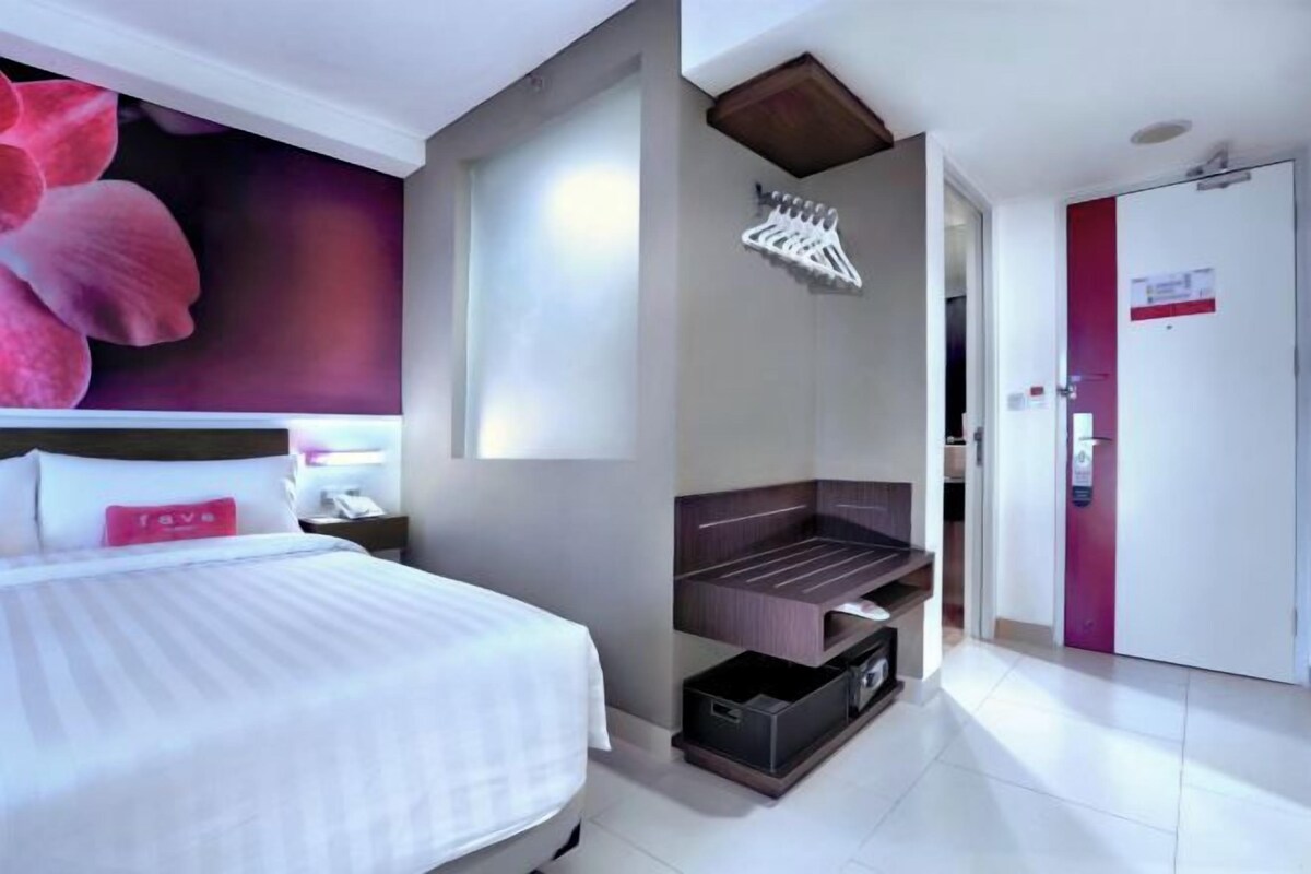 Quarto de hotel moderno com cama grande, um banco embutido, um suporte fixado na parede e uma porta com uma placa.
