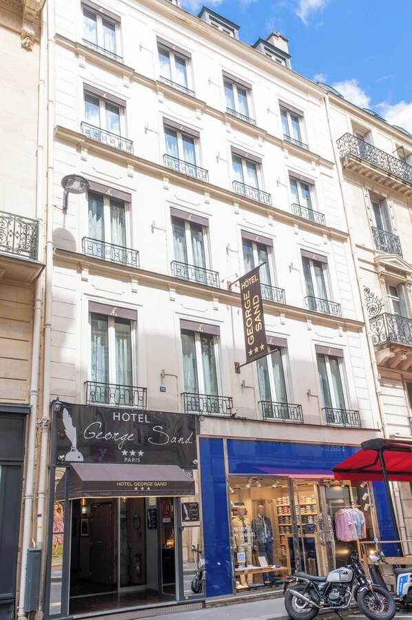 Fachada de hotel com uma placa indicando "HOTEL GEORGE SAND PARIS", uma vitrine com manequins e uma motocicleta estacionada na frente.