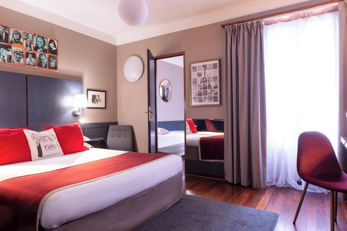 Quarto de hotel com uma cama grande, uma mesa, uma cadeira, um espelho e uma janela com cortinas.