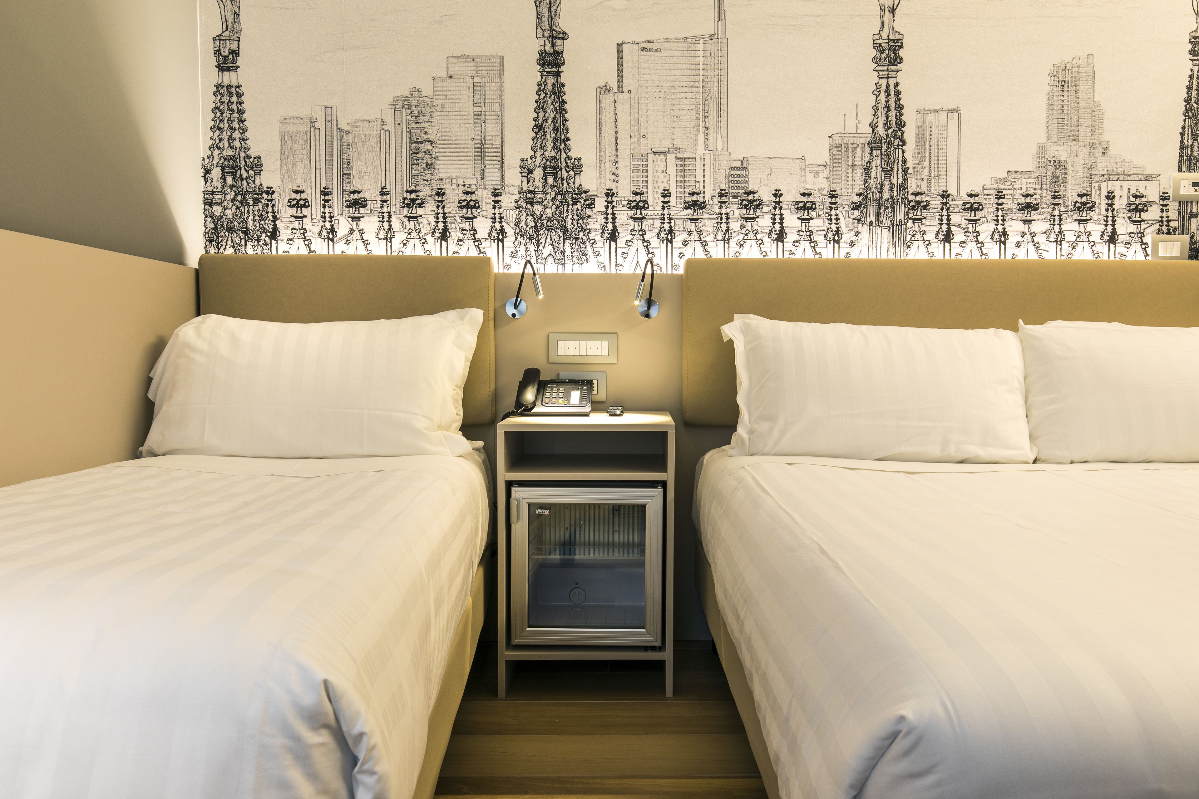 Quarto de hotel com duas camas, uma mesa de cabeceira com telefone e um mural com vista para a cidade.