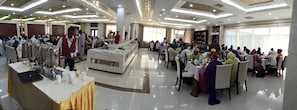 Uma ampla sala de jantar com várias mesas e cadeiras, um buffet central e um teto bem iluminado com detalhes decorativos.
