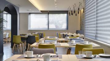 Uma área de refeições moderna com mesas preparadas para o café da manhã, apresentando uma variedade de cadeiras e uma grande janela com persianas.
