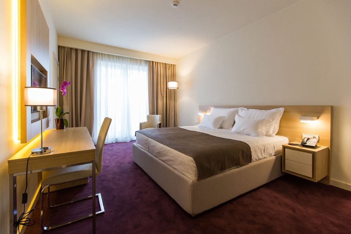 Quarto de hotel com cama, mesa de cabeceira, luminária, escrivaninha, cadeira e uma janela grande com cortinas.