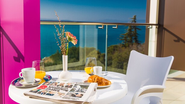 Mesa de café da manhã com jornal, croissant, suco de laranja e xícaras de café, com vista panorâmica.