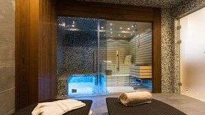 Uma sala de spa com duas mesas de massagem, uma banheira de hidromassagem em vidro e um banheiro moderno com área de chuveiro revestida em azulejo.