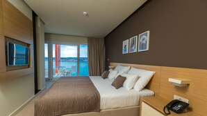Quarto de hotel moderno com uma cama grande, telefone ao lado da cama e vista para o mar através de uma porta de vidro deslizante.