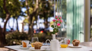 Uma mesa com um jarro branco, uma xícara com o logotipo da marca de café, um muffin, um copo de suco de laranja e um vaso com flores cor-de-rosa.