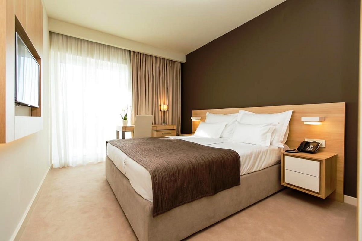 Quarto de hotel com uma cama grande, mesas de cabeceira, escrivaninha e televisão na parede.